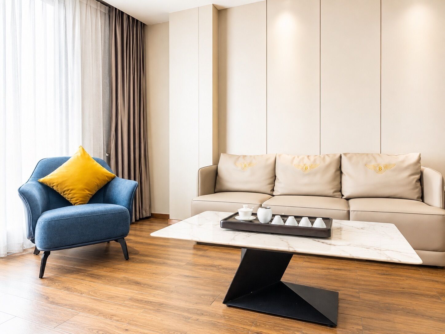 Uma sala de estar moderna com uma poltrona azul, um sofá bege e uma mesa de centro com uma bandeja de bules brancos.