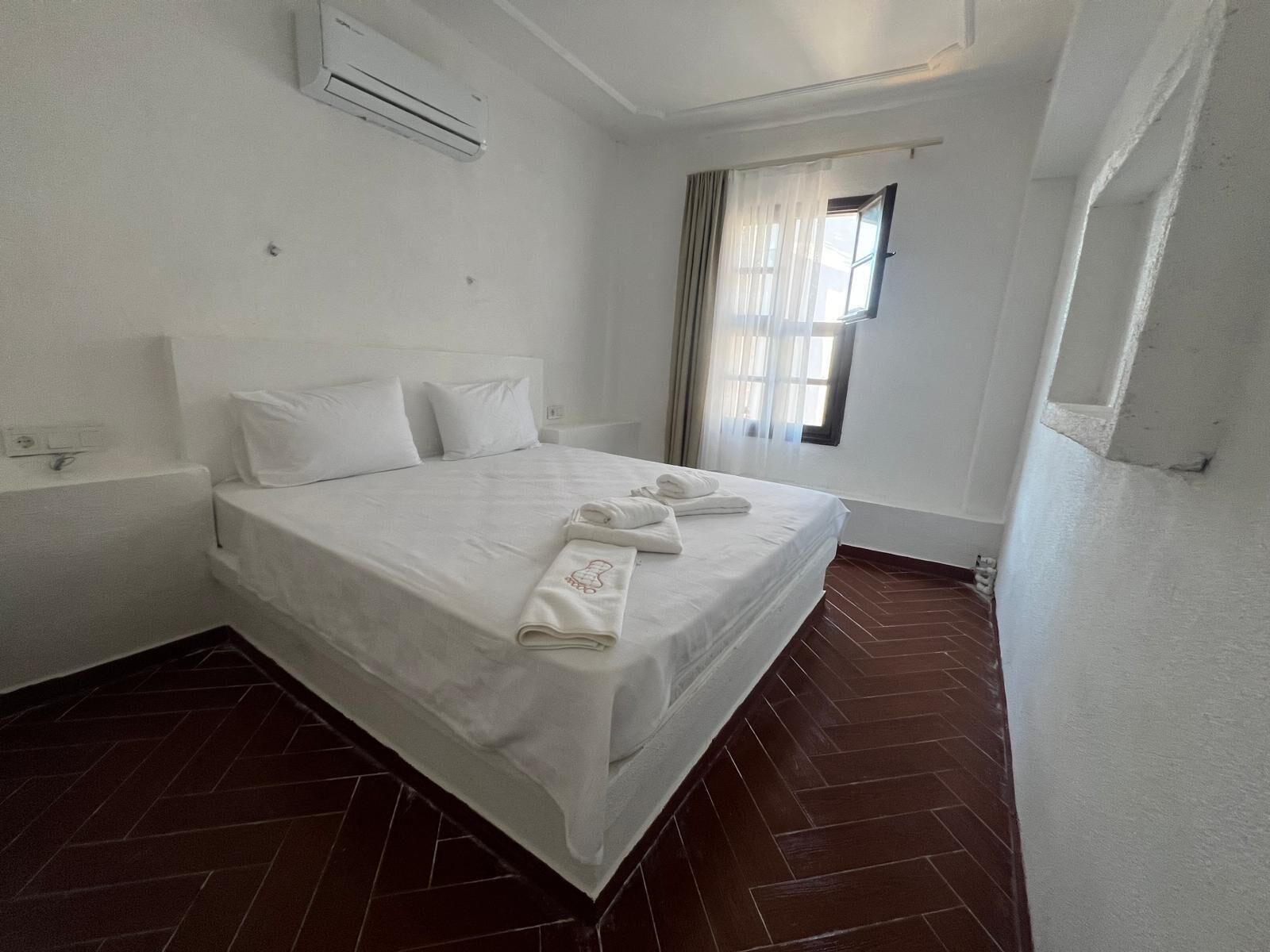 Quarto de hotel com cama, paredes brancas, janela com cortinas e piso de azulejos.