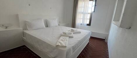 Quarto de hotel com cama, paredes brancas, janela com cortinas e piso de azulejos.
