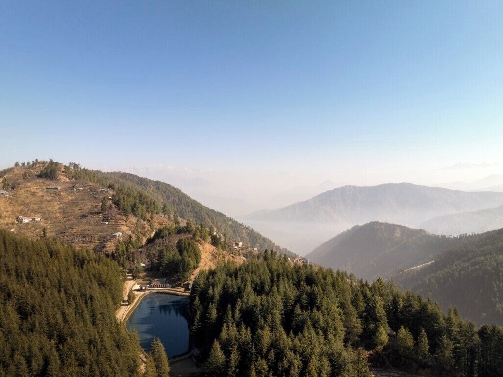 Paisagem montanhosa com um lago, áreas arborizadas e montanhas ao longe.