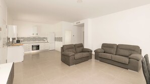 Uma sala de estar espaçosa com dois sofás e uma cozinha ao fundo, tudo em tons de branco.