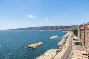Paisagem costeira urbana com céu azul límpido, um calçadão à beira-mar e vista distante de uma cidade em colinas.