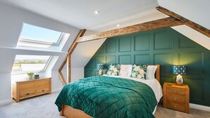 Quarto com teto inclinado, janelas de claraboia, cama com colcha verde, mesinhas de cabeceira de madeira e abajur.