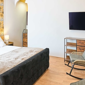 Um quarto com cama, mesa de cabeceira, cômoda, cadeira e televisão na parede.