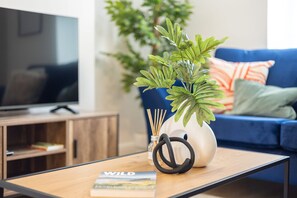 Uma sala de estar moderna com uma mesa de centro de madeira, um vaso branco com plantas verdes, uma revista intitulada 'WILD', uma televisão e um sofá azul com almofadas.