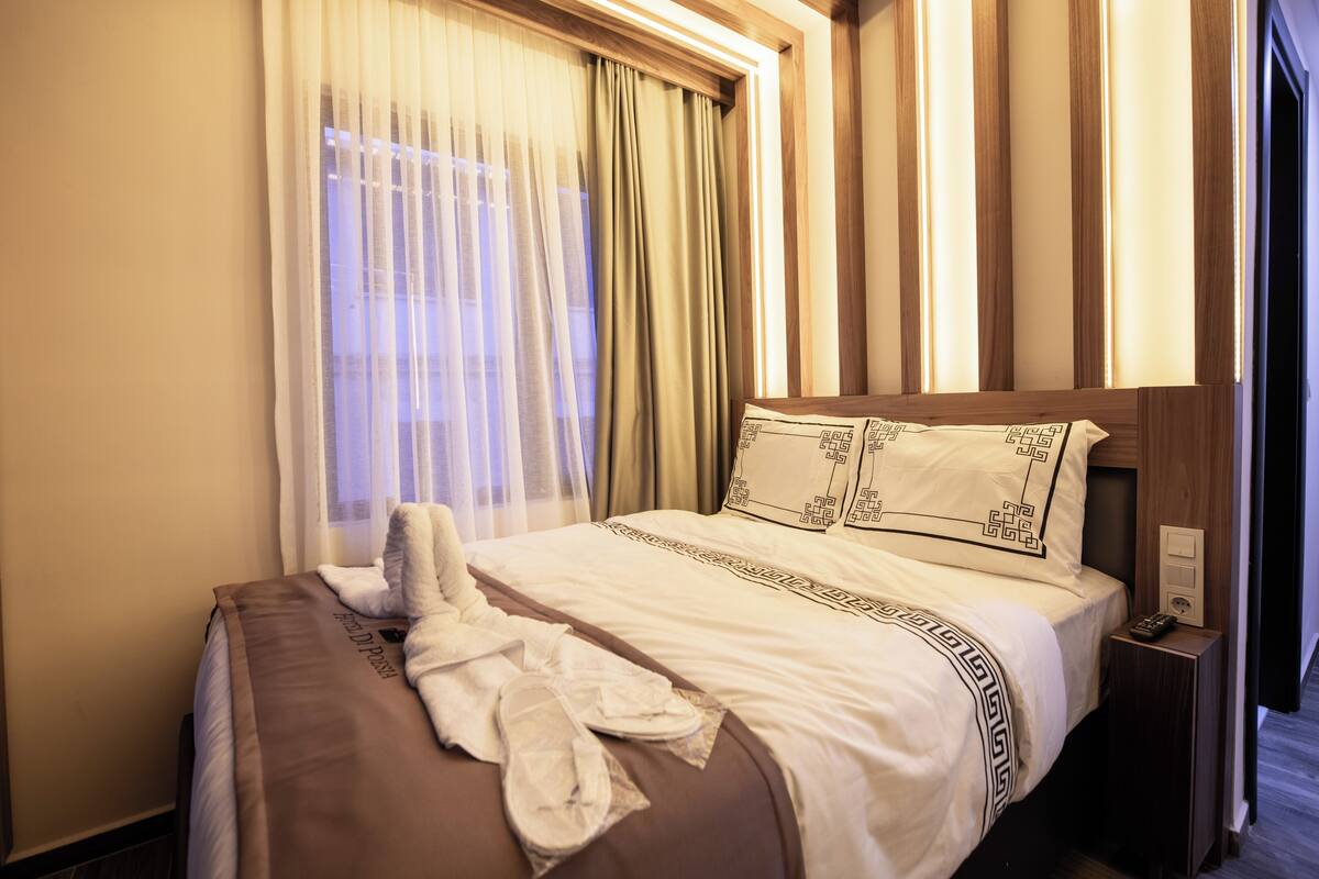 Quarto de hotel com cama bem arrumada, mesa de cabeceira com controle remoto e janela com cortinas translúcidas.