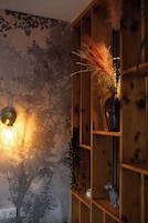 Quarto com um mural na parede, prateleira de madeira com um vaso de plantas secas, luminária fixada na parede e uma pequena estatueta de gato.