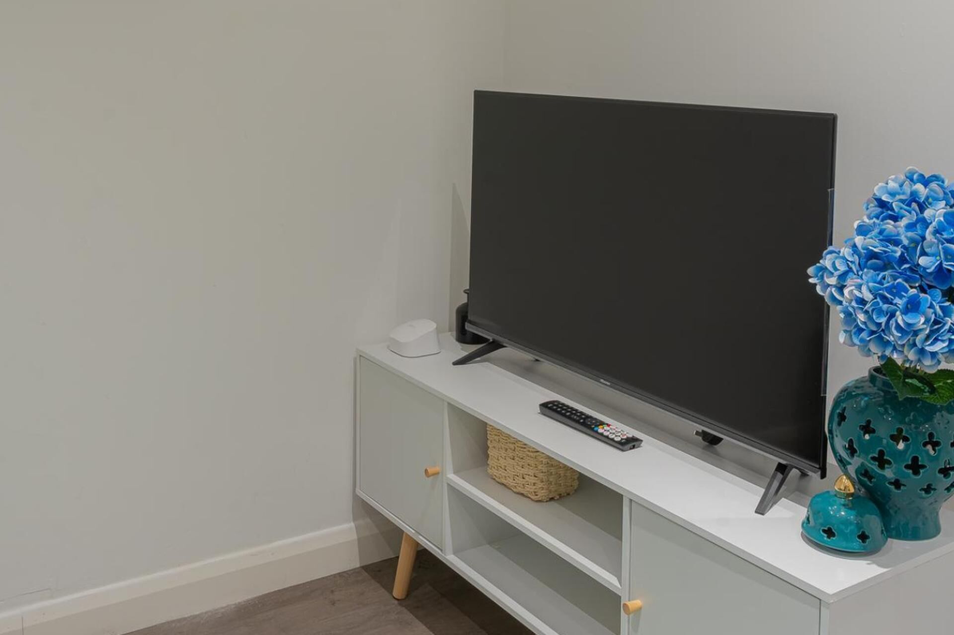 Um móvel branco para televisão com uma TV de tela plana, um controle remoto e um vaso com flores azuis.
