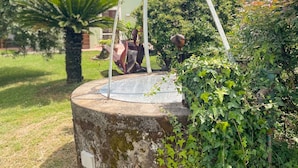 Uma atração aquática rústica ao ar livre, com uma bacia de pedra e arco de metal, cercada por vegetação exuberante e uma casa ao fundo.
