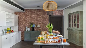 Uma área de refeições com bancada de mármore, variedade de comidas e bebidas, luminária pendente decorativa e papel de parede estampado.