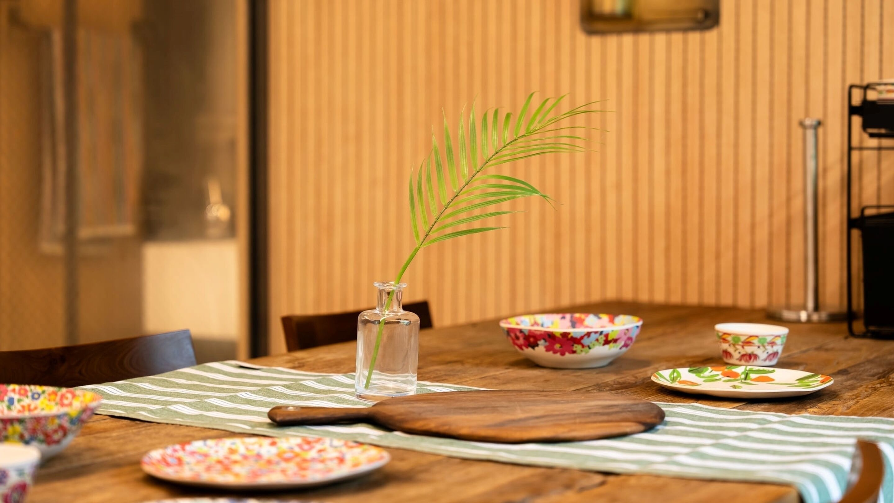 Uma mesa de jantar de madeira com toalha listrada, um vaso de vidro transparente com uma única folha verde, e tigelas e pratos com padrão floral colorido.