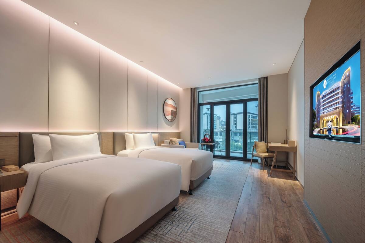 Quarto de hotel moderno com duas camas grandes, uma televisão de tela plana e uma janela ampla com vista para a cidade.