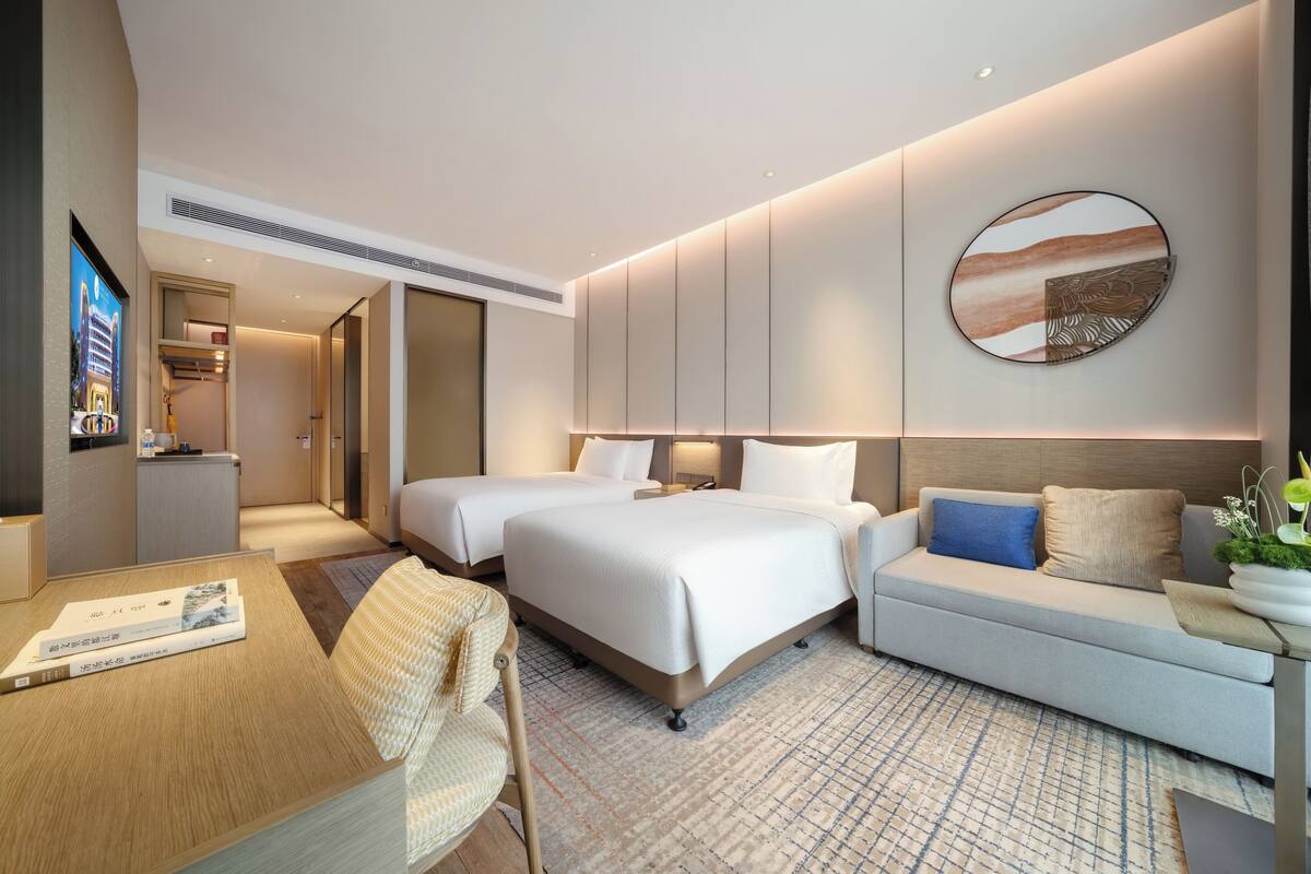 Quarto de hotel moderno com duas camas, um sofá, uma escrivaninha e um espelho grande.