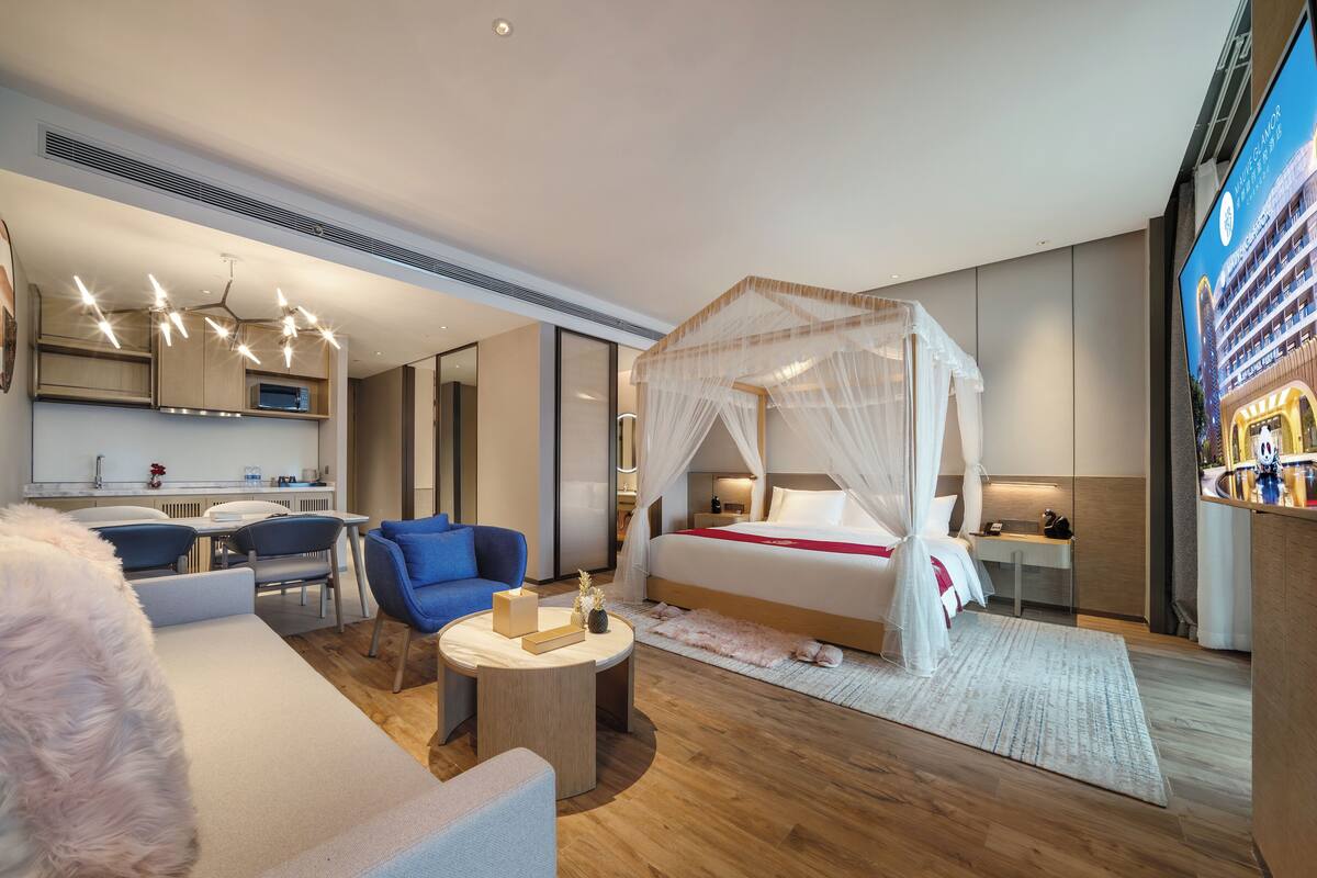 Quarto de hotel moderno com uma cama grande, área de estar com sofá e poltronas, pequena cozinha e uma janela ampla.