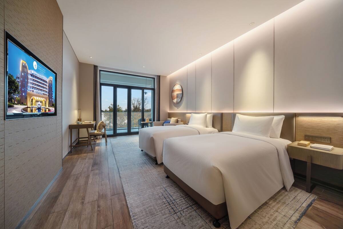 Quarto de hotel moderno com duas camas, televisão na parede e uma janela grande.