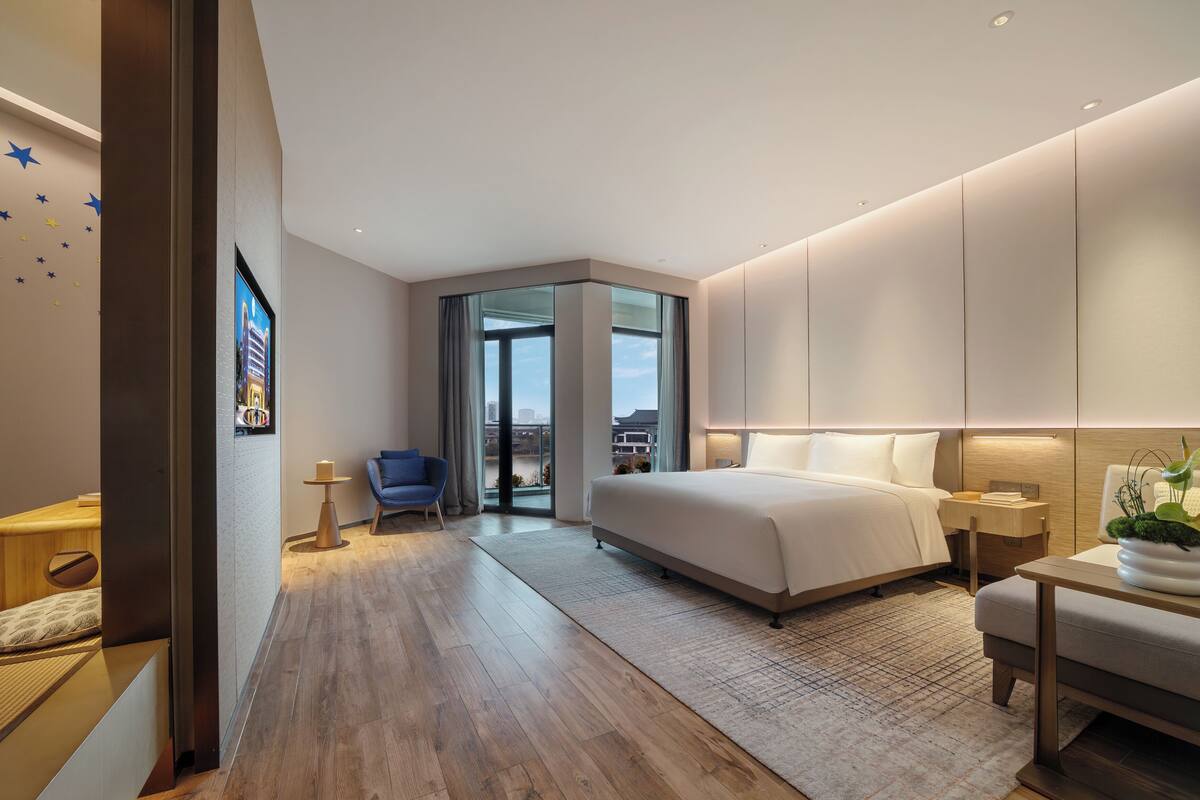 Quarto de hotel moderno com uma cama grande, um espaço de estar com uma poltrona, uma mesinha e vista para a cidade através de grandes janelas.