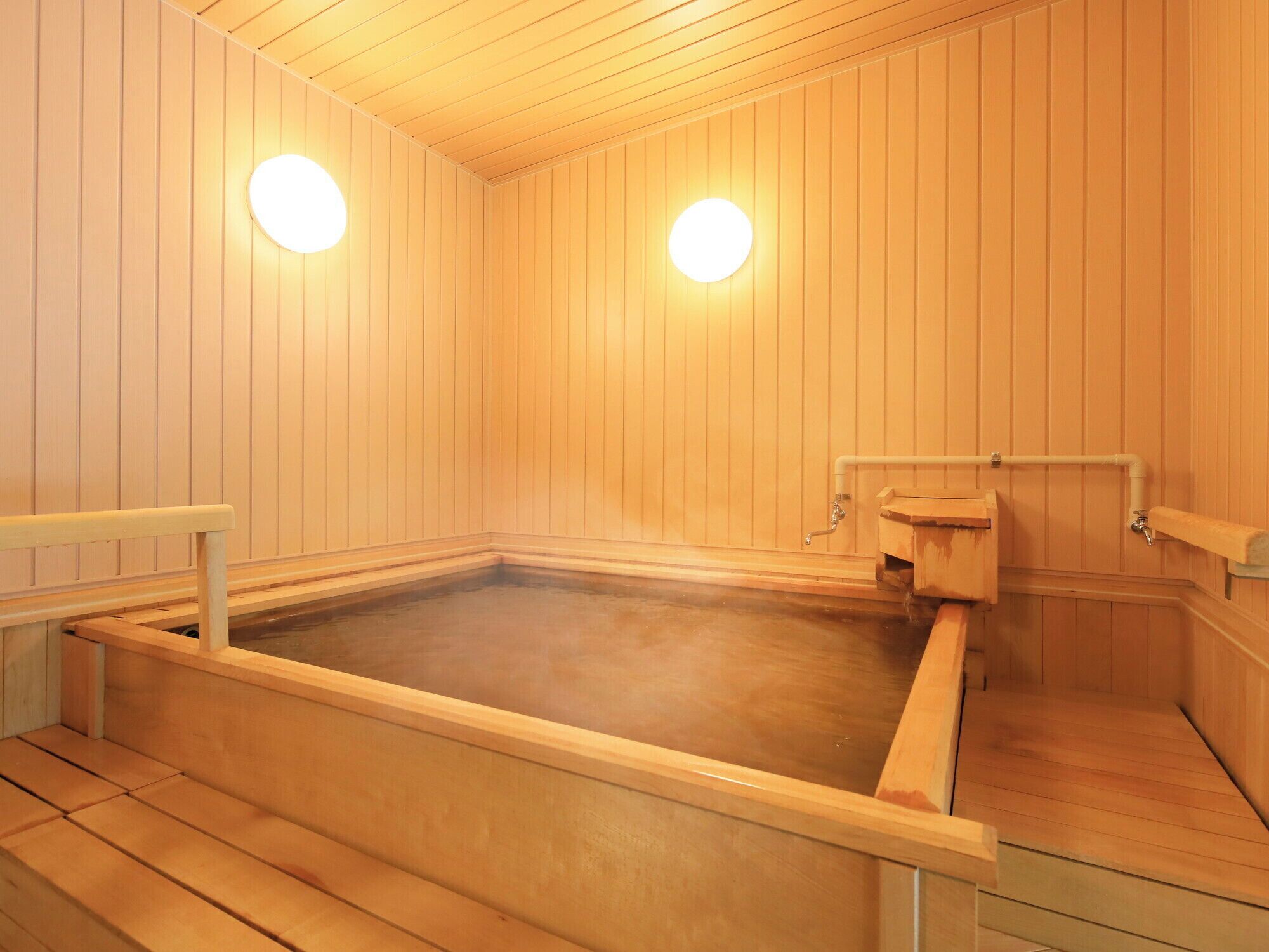 Uma sauna de madeira com um banco, uma pequena estrutura de madeira com uma torneira e duas luminárias redondas na parede.