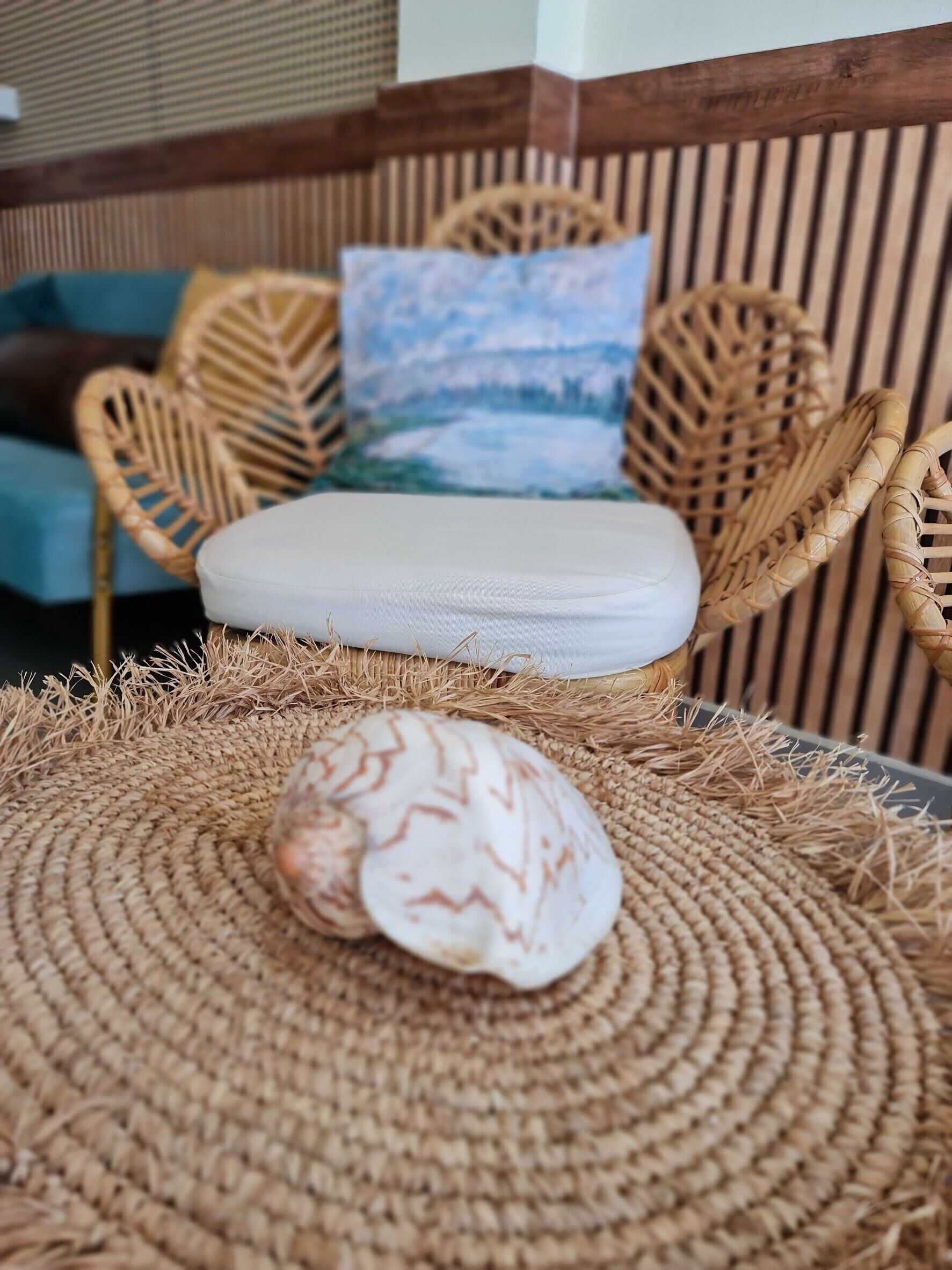 Uma cadeira de vime com almofada, um tapete de palha com conchas e um sofá azul com uma pintura de paisagem em um quarto com lambris de madeira.