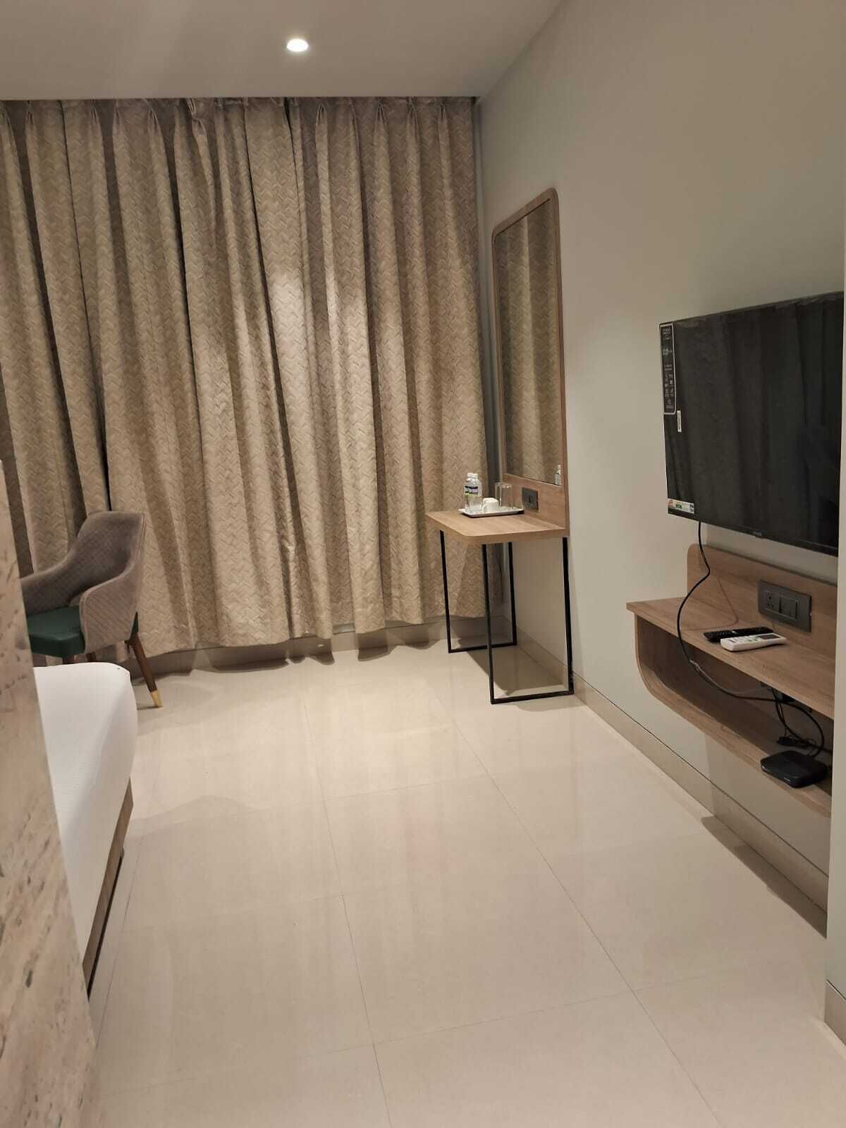 Quarto de hotel com cama, escrivaninha com cadeira, televisão e uma janela grande com cortinas.