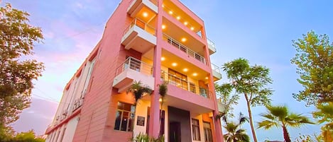 Um hotel de vários andares com fachada rosa, varandas em cada pavimento e uma entrada central com degraus.