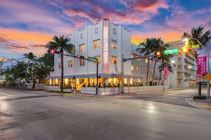 Hotel com arquitetura clássica, exibindo uma marquise em destaque, situado no cruzamento de uma rua com palmeiras e céu ao pôr do sol.