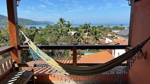 Uma rede numa varanda com vista para a costa, com casas e palmeiras.