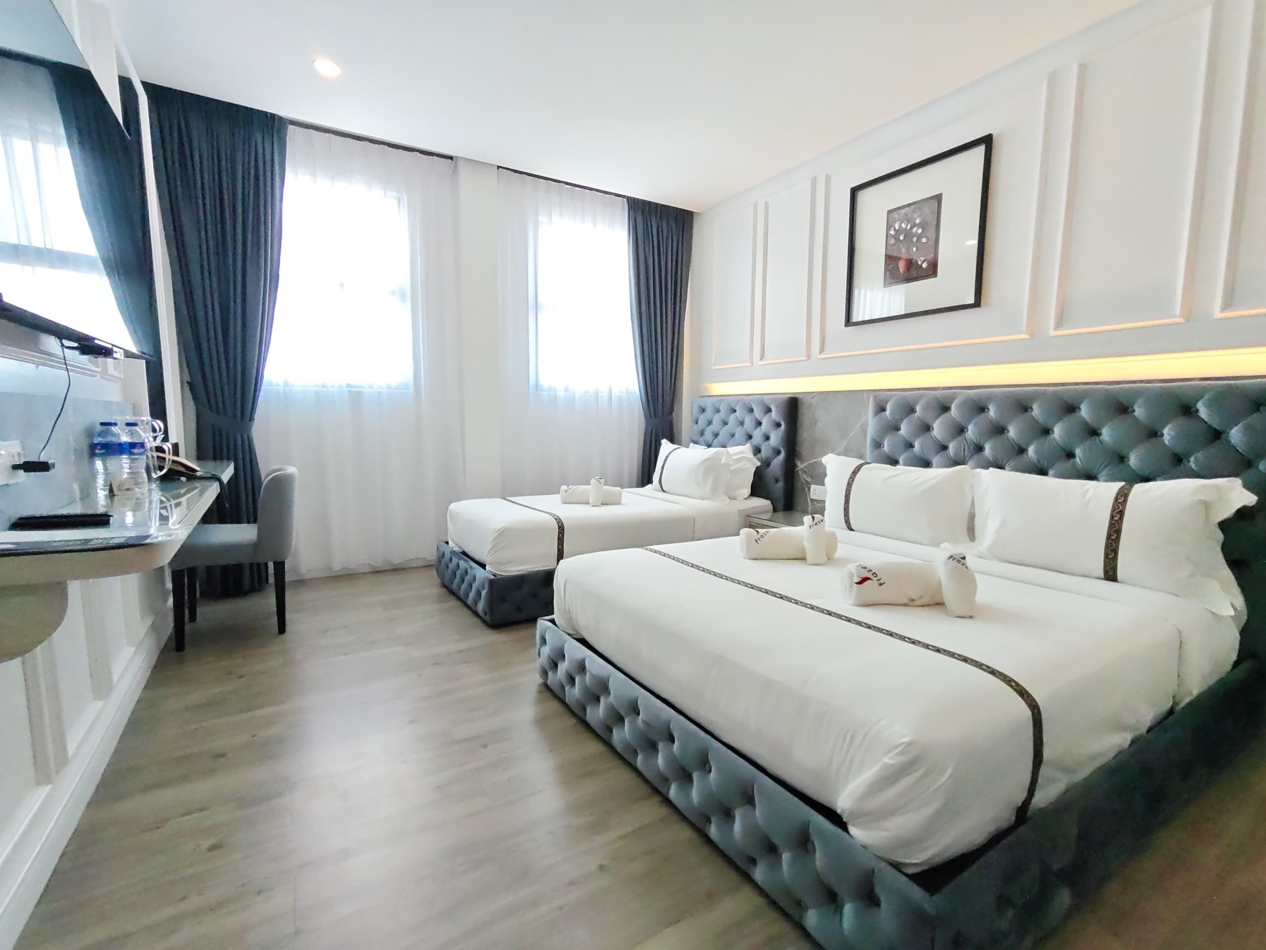 Quarto de hotel com uma cama grande, uma cama menor, uma escrivaninha com cadeira e uma janela com cortinas.