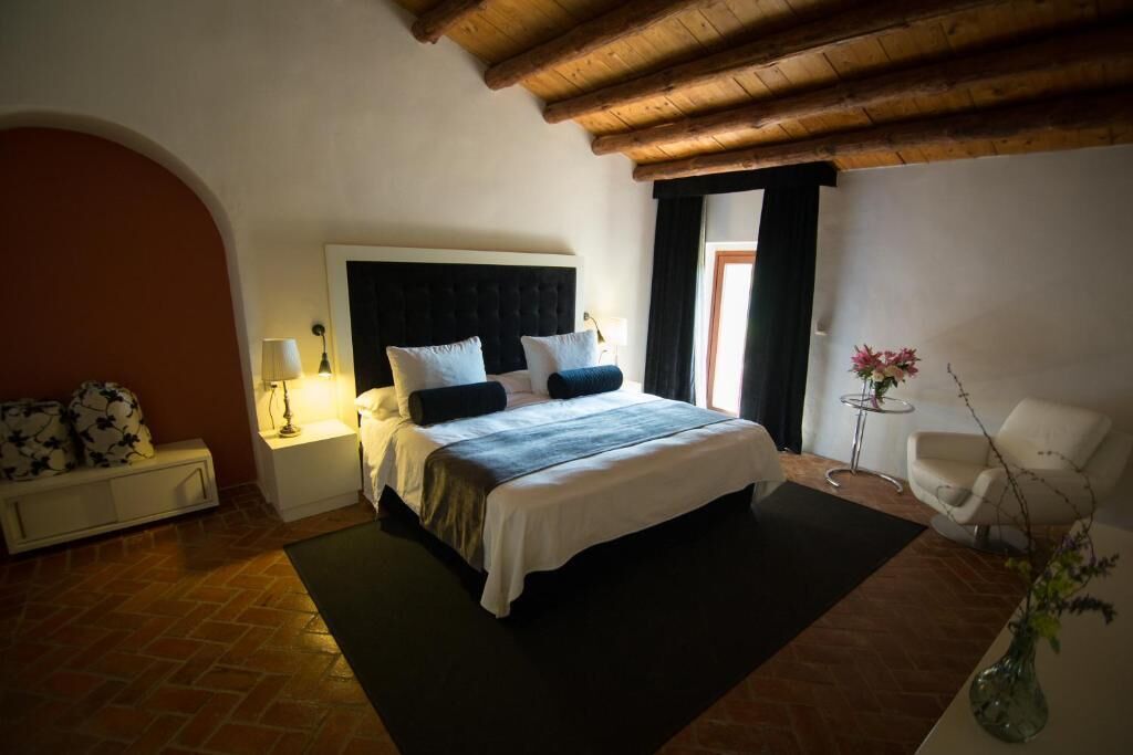 Quarto com uma cama grande, mesa de cabeceira com abajur, cadeira e vaso com flores.