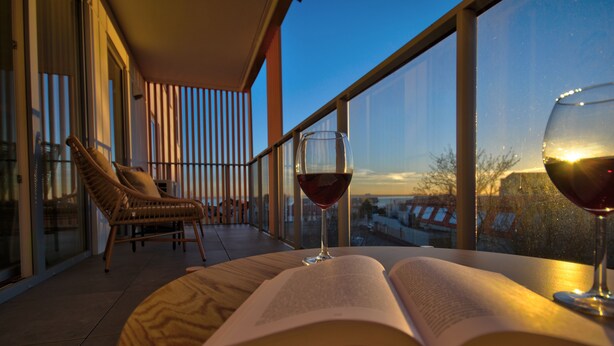 Um livro aberto sobre uma mesa de madeira com dois copos de vinho numa varanda com vista para uma paisagem urbana.