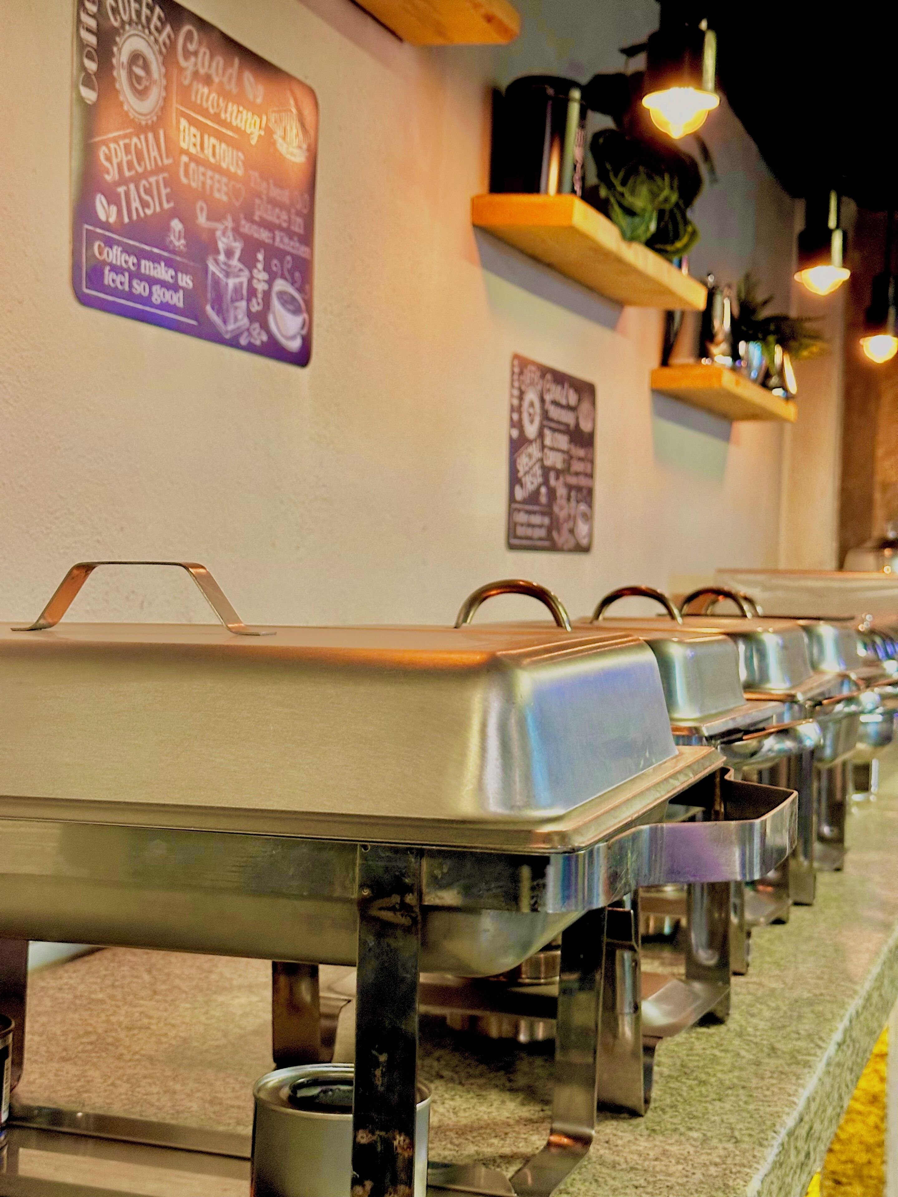 Buffet com bandejas de aço inoxidável e parede com sinalização sobre café.