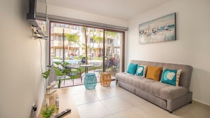 Uma sala de estar moderna com um sofá cinzento, almofadas verde-água e laranja, uma pintura de veleiros e vista para uma piscina e edifícios.