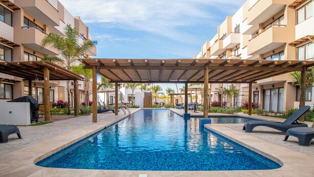 Piscina com espreguiçadeiras e um pérgola, cercada por edifícios com varandas.