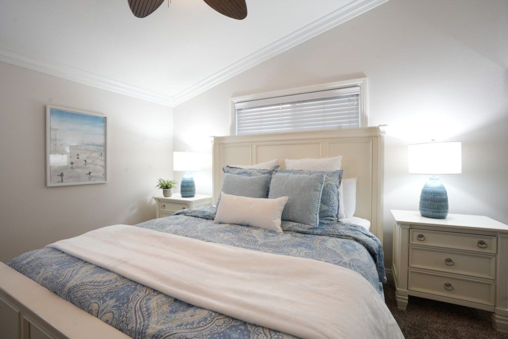 Um quarto com cama, mesas de cabeceira, abajur, janela com persianas e um quadro na parede.