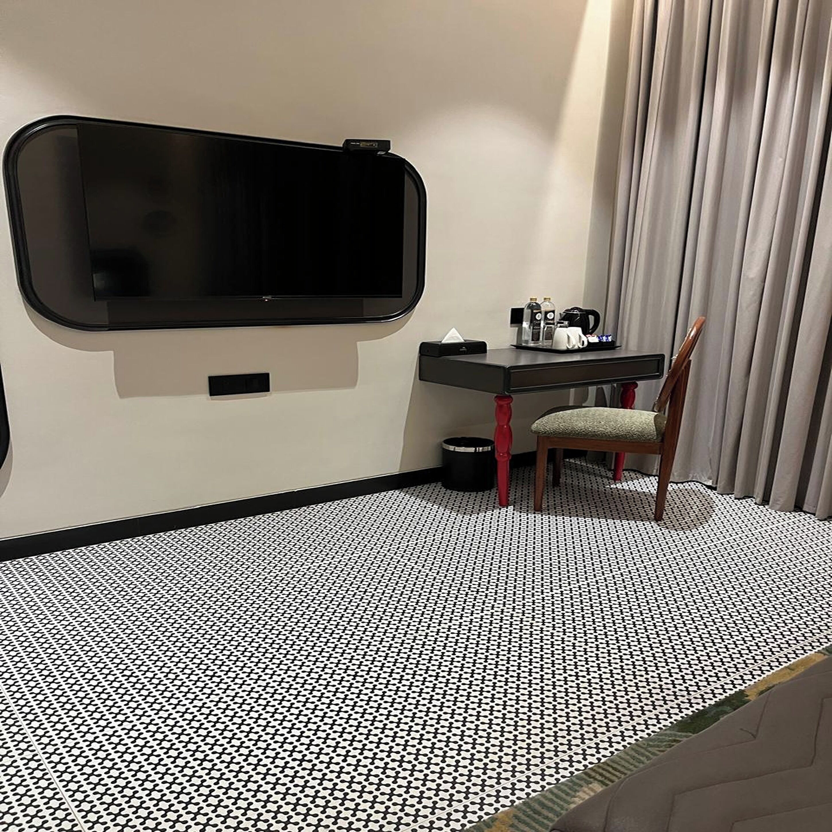 Quarto de hotel com chão padronado em preto e branco, uma pequena mesa com cadeira, TV de tela plana na parede e uma janela com cortinas.