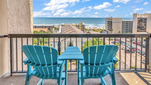 Uma varanda com duas cadeiras Adirondack turquesa e uma mesa, com vista para uma praia e um estacionamento.