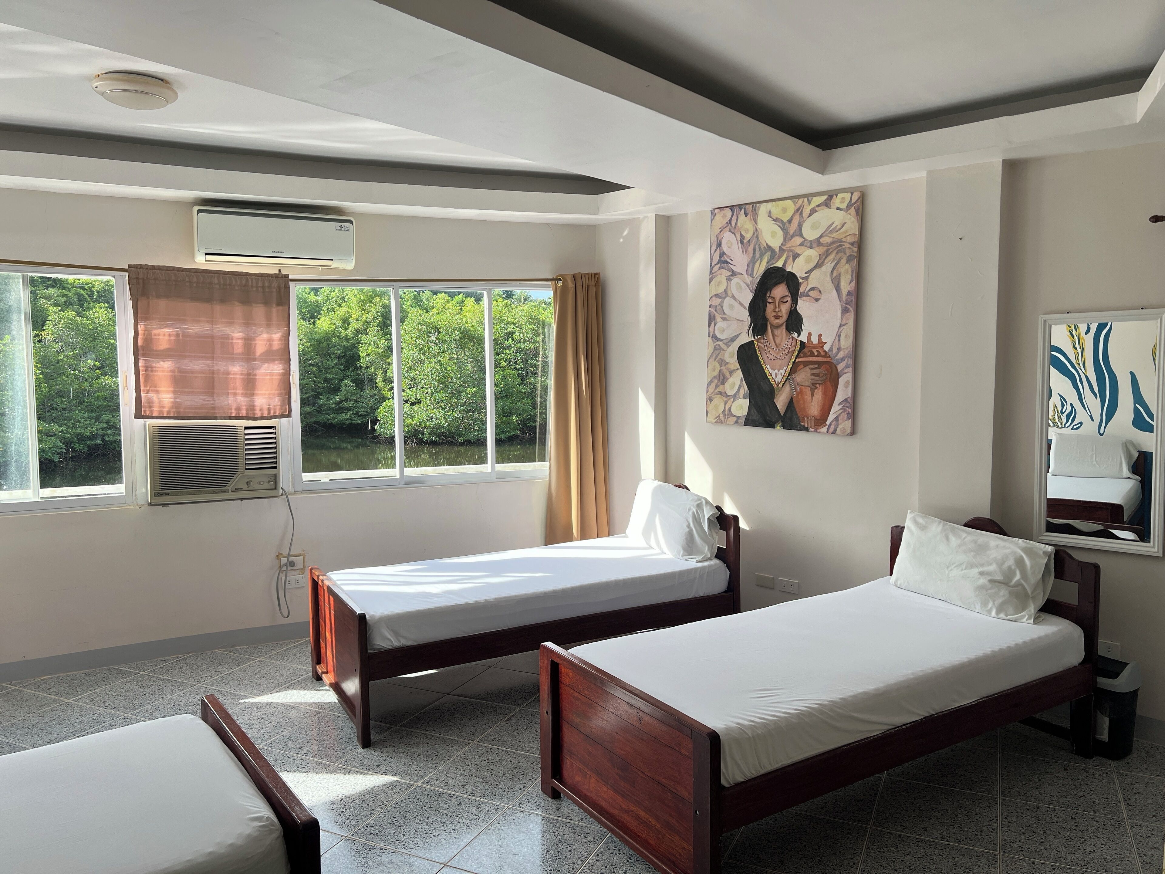 Quarto de hotel com duas camas de solteiro, uma janela grande com cortinas, um aparelho de ar condicionado e um quadro na parede.
