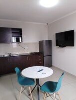 Cozinha moderna com armários de madeira escura, micro-ondas, fogão, geladeira e uma televisão de tela plana na parede. Há uma mesa redonda com duas cadeiras verde-água e uma bancada branca.
