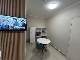 Uma cozinha pequena e compacta com geladeira branca, micro-ondas e armário branco. Há uma mesa redonda com duas cadeiras e uma televisão na parede.