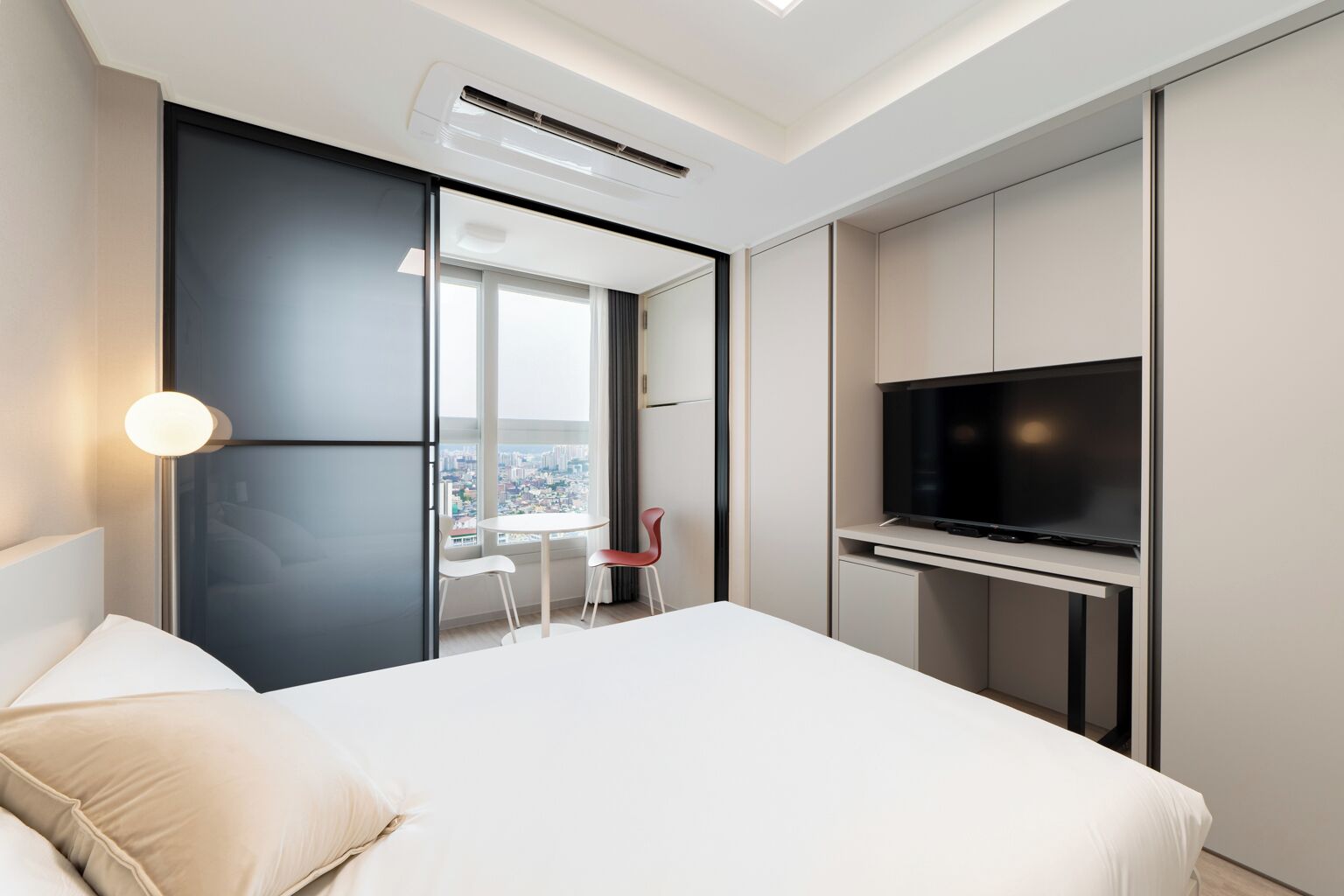 Quarto de hotel moderno com uma cama, televisão, uma pequena mesa com cadeiras e vista através de uma grande janela.