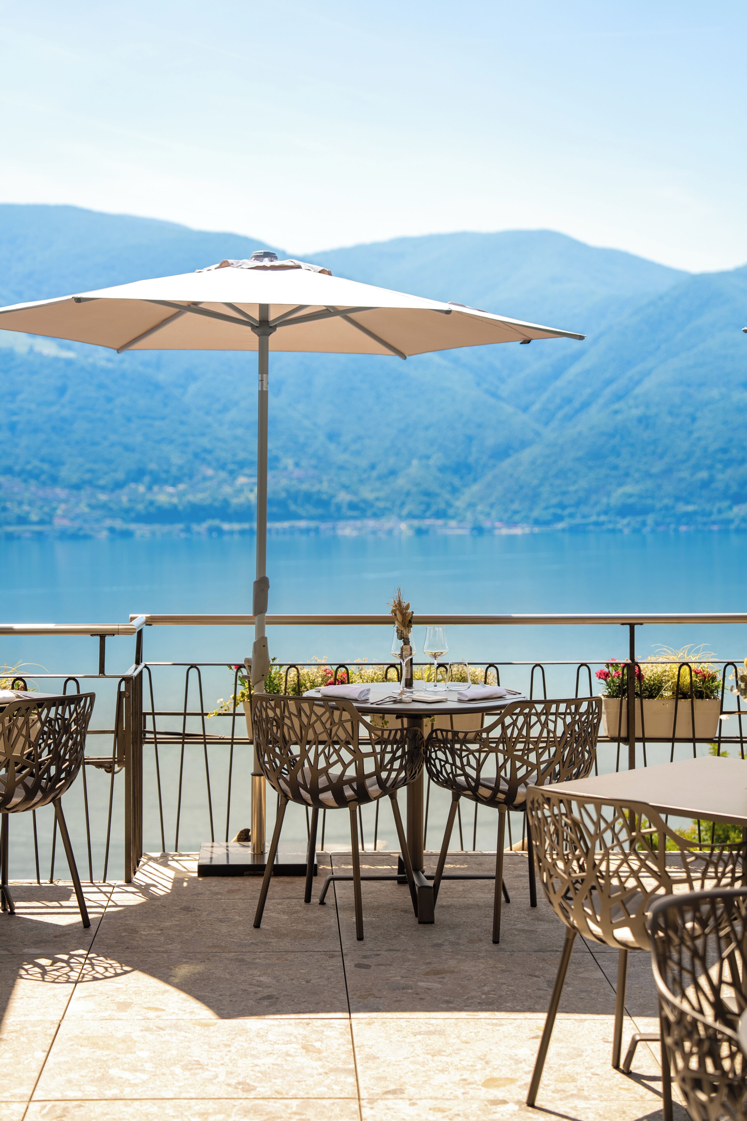 Uma área de refeições ao ar livre com mesa, cadeiras, um grande guarda-sol e vista para montanhas e um lago.