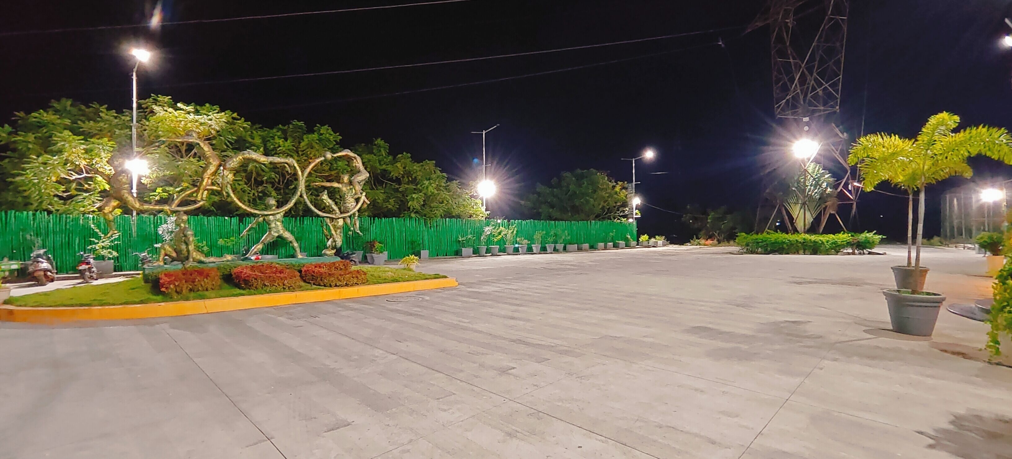 Um pátio pavimentado com uma escultura de metal decorativa, sebes verdes e plantas em vasos.