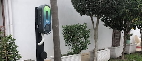 Uma área de jardim com um posto de carregamento para veículo elétrico preto, plantas em vasos e um caminho pavimentado com azulejos.