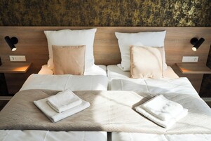 Uma cama de casal com duas almofadas, uma toalha dobrada e cabeceira com luzes fixadas na parede.