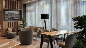 Quarto de hotel moderno com uma grande janela, área de refeições com mesa e cadeiras, e um espaço de estar com sofás e um abajur.