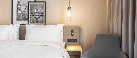 Quarto de hotel bem organizado com uma cama, uma poltrona, mesa de cabeceira com abajur e duas fotografias emolduradas na parede.