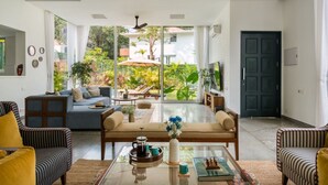 Sala de estar ampla com sofá, poltronas e mesa de centro. Vista para um jardim através de janelas grandes.