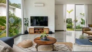 Uma sala de estar moderna com uma televisão grande, assentos confortáveis, uma mesa de centro e vista para o verde lá fora.