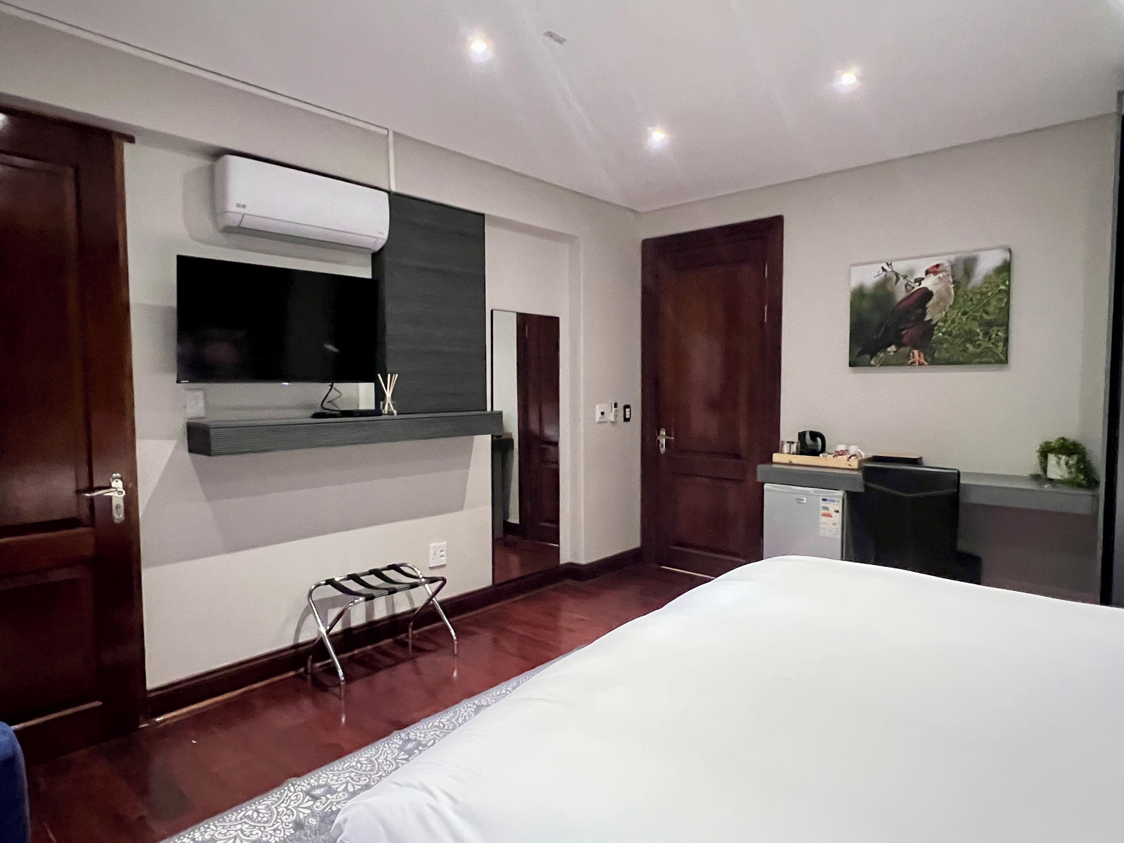 Quarto de hotel com cama, escrivaninha, cadeira, televisão, guarda-roupa e porta.