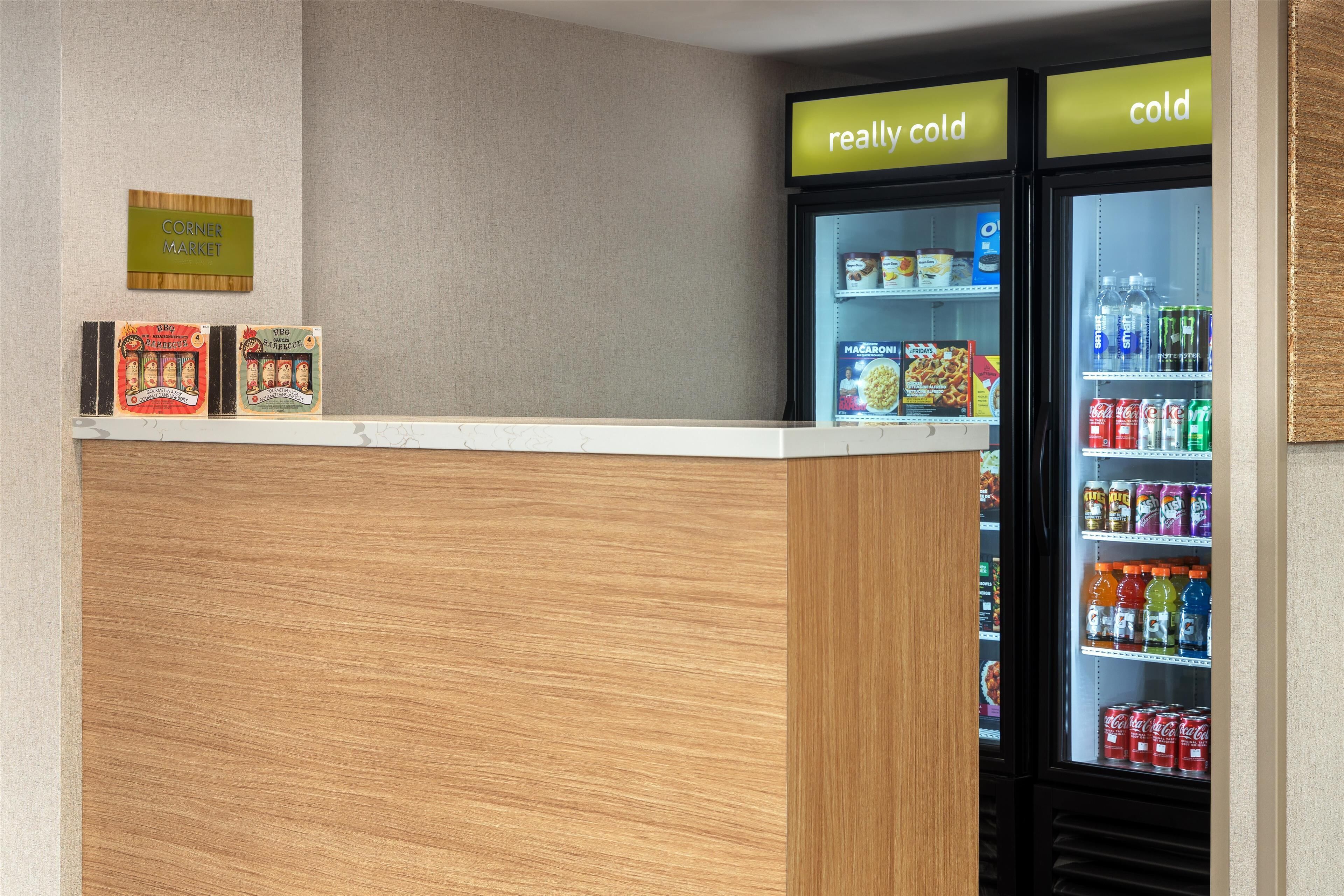 Uma área de recepção com balcão de madeira, um letreiro 'Corner Market' e dois dispensadores de bebidas com a etiqueta 'bem gelado' e 'gelado'.
