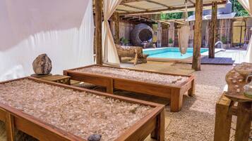 Uma área de spa com banheiras de madeira cheias de sal, uma rede pendurada e uma piscina ao fundo.